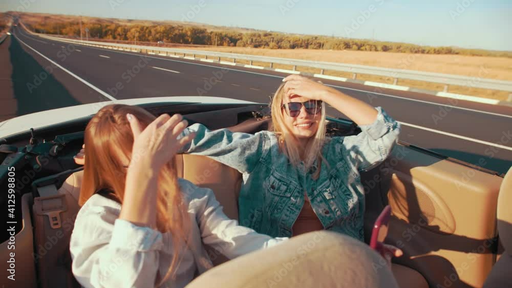 Women in sunglasses take selfies while posing in a convertible, happy ...