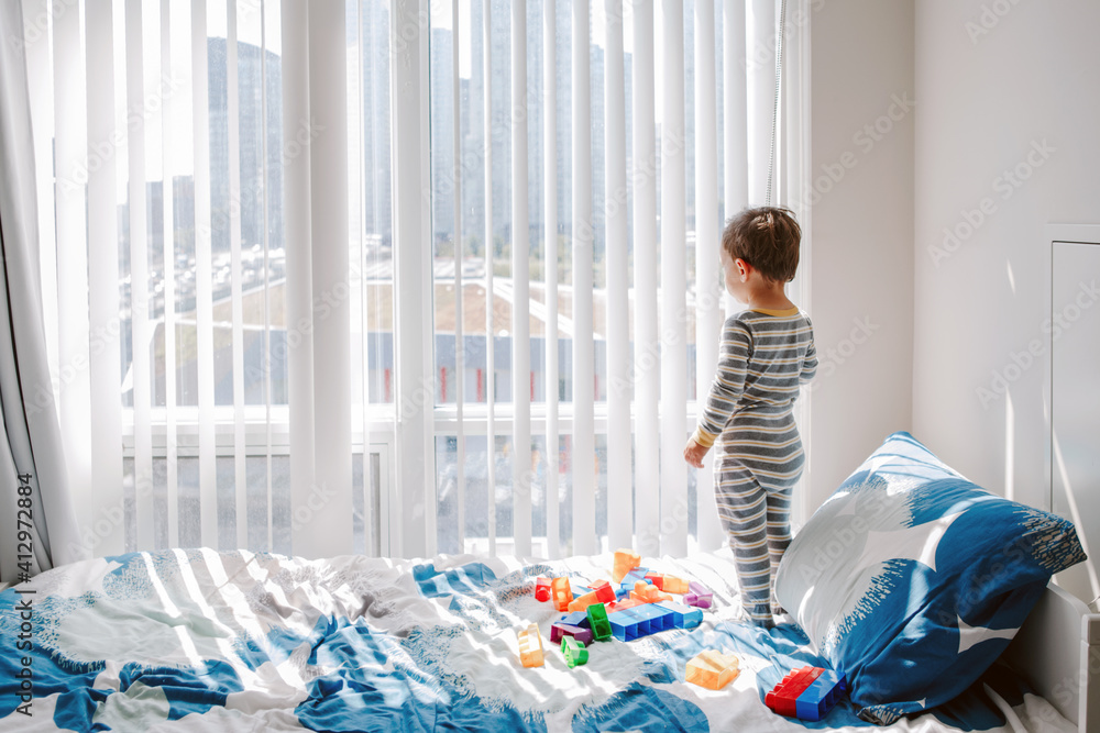 Cute little boy toddler standing on bed in room and looking into window ...