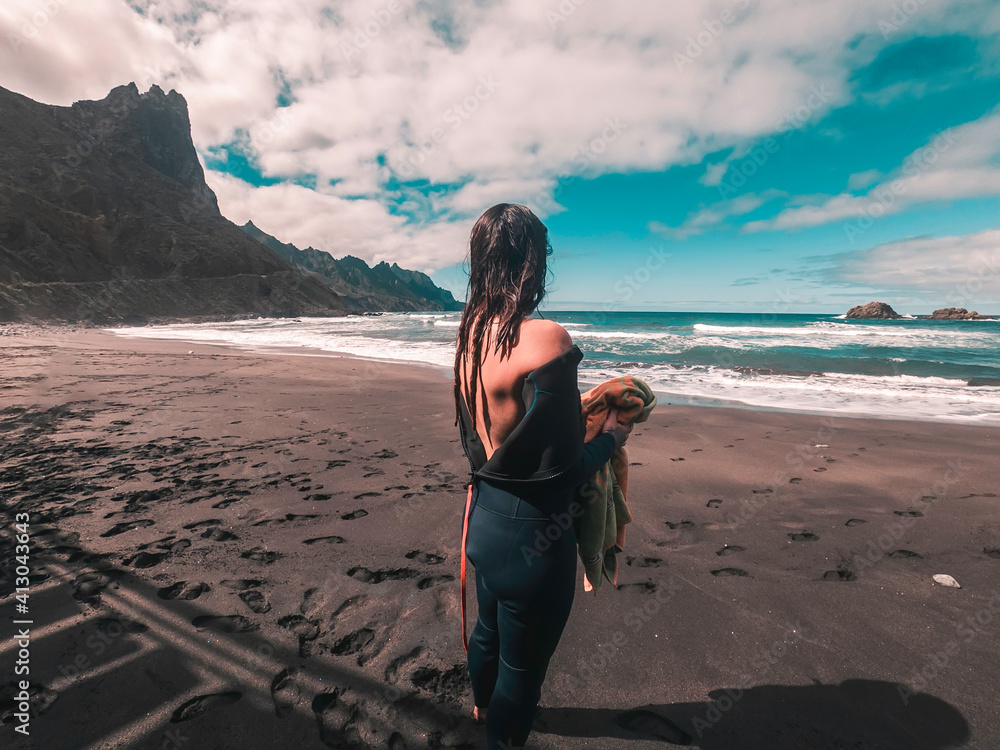 Young Surfer Woman Taking Off A Wet Neoprene Wetsuit On The Black Sand