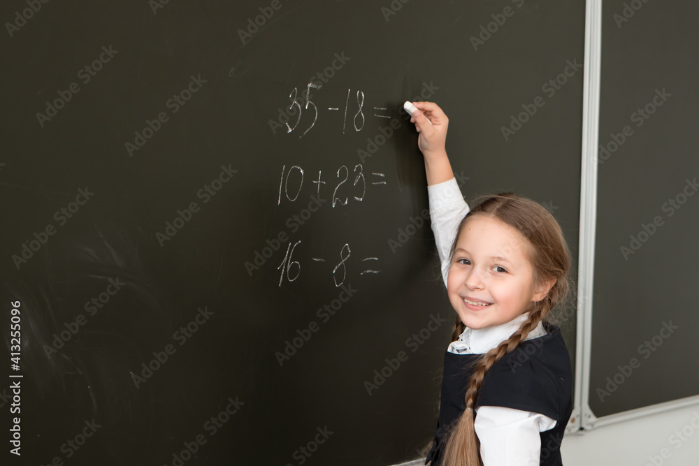 smiling little girl solve math task in front of blackboard in classroom ...