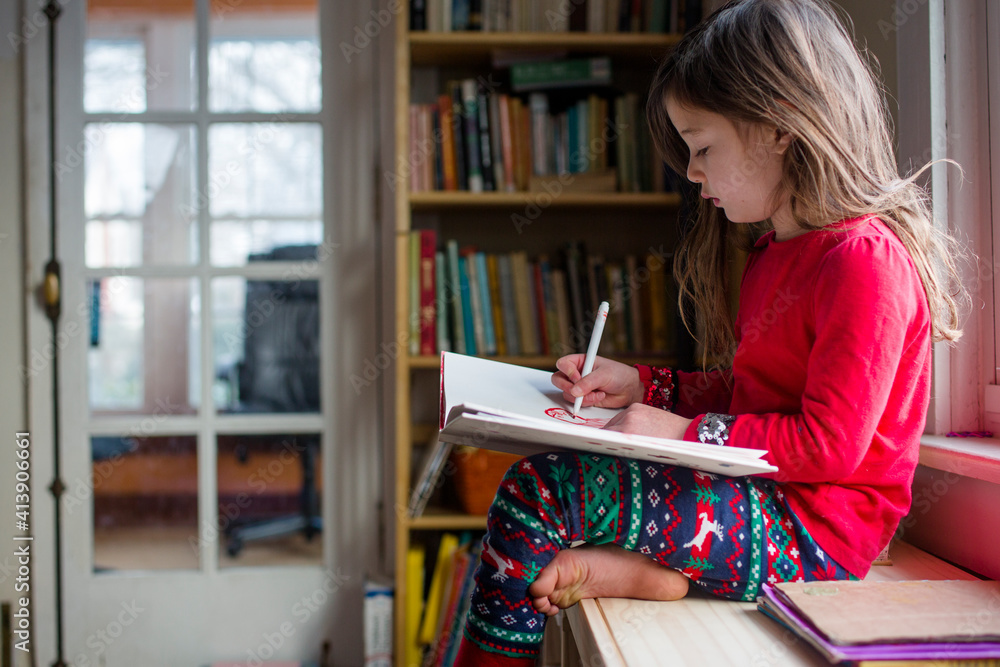 side view of a little girl drawing pictures in her pajamas by window ...