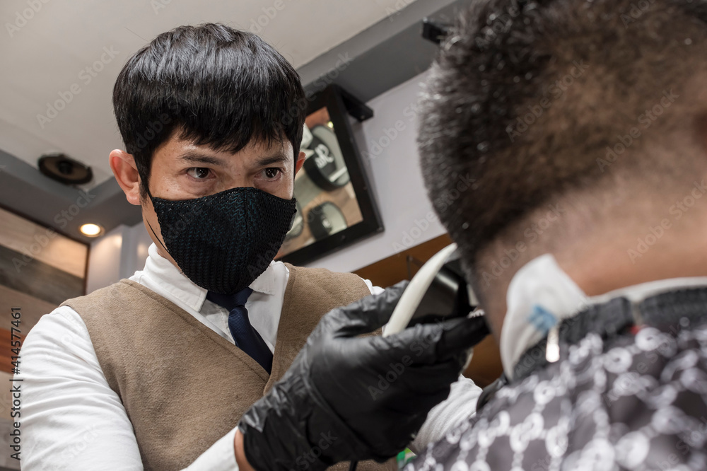 An asian barber wearing a face mask trims and fades the back of a ...