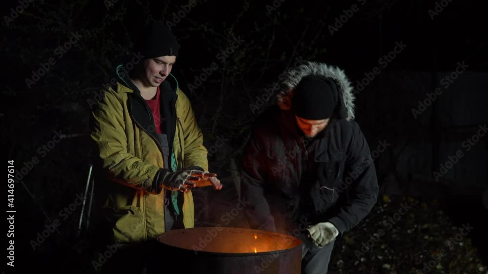 Two homeless young men bask in the fire. Men stand near a barrel of ...