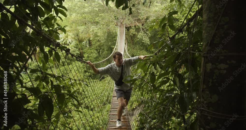 Front static view of a man walking across a shaky rope jungle bridge in ...