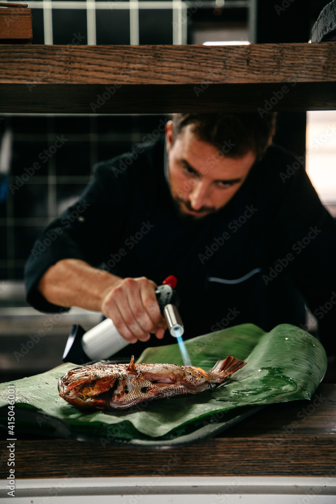 Concentrated male chef cooking fried fish with gas torch while working ...