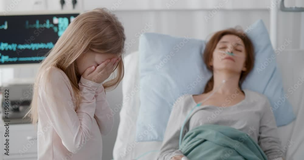 Little girl crying standing near sick mother sleeping in hospital bed ...