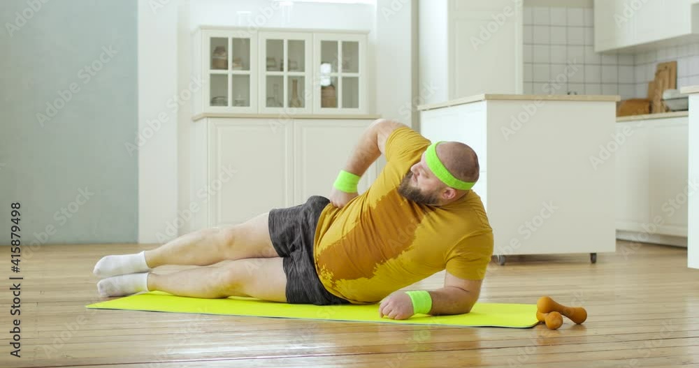 Sweaty fat man in yellow wet sportswear is doing legs lifting exercises ...