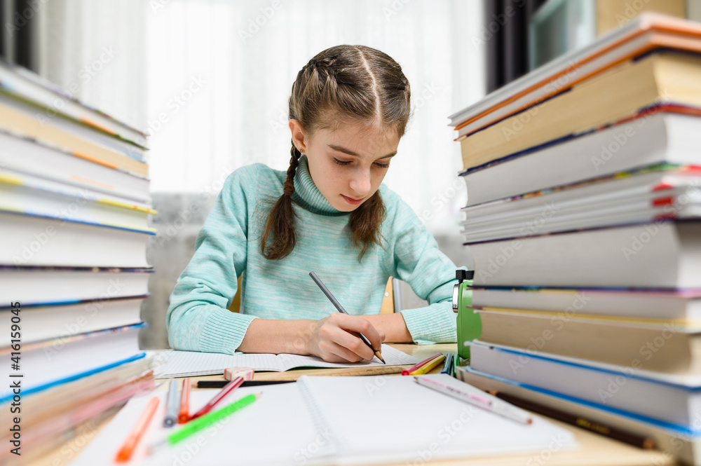 Schoolgirl doing homework at her desk. Next to the girl are tall stacks ...