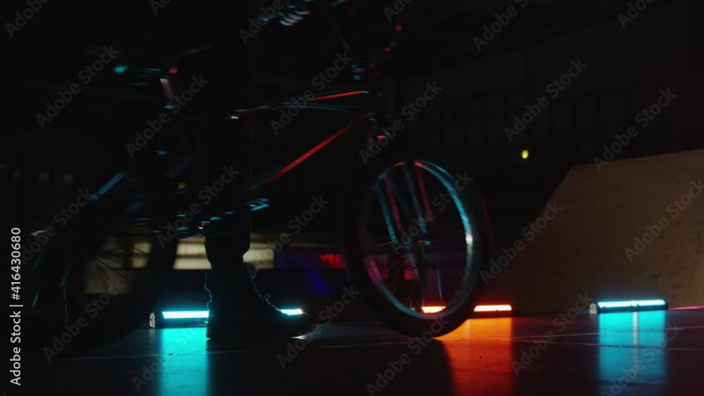 BMX rider walks bike across indoor ramps with neon lights on the ground ...