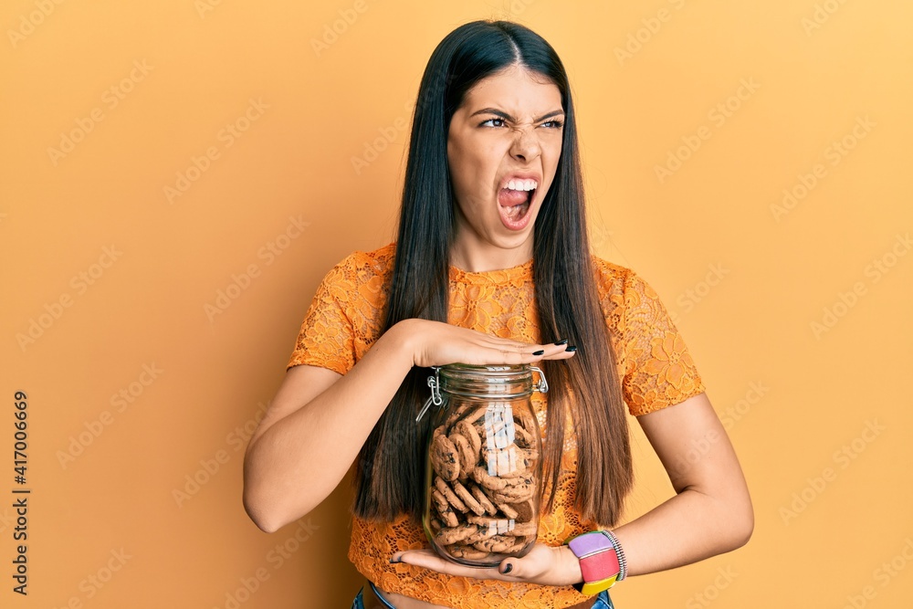Young hispanic woman holding jar with chocolate chips cookies angry and ...