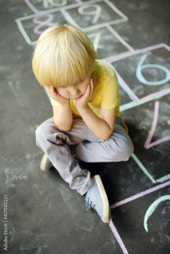 A sad little boy sitting on the asphalt with classics painted on it ...