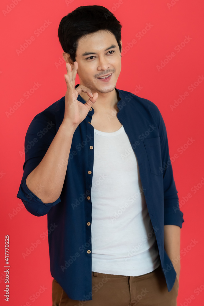 Young Asian man shows a hand sign and smiles while standing on a red ...