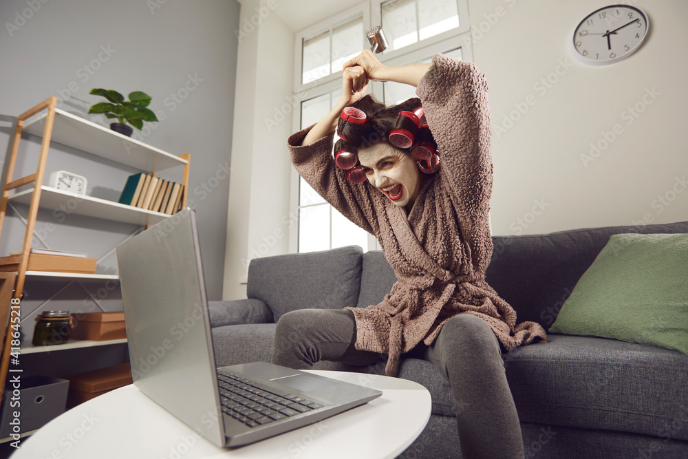 Funny furious young woman in hair curlers breaking computer with hammer ...