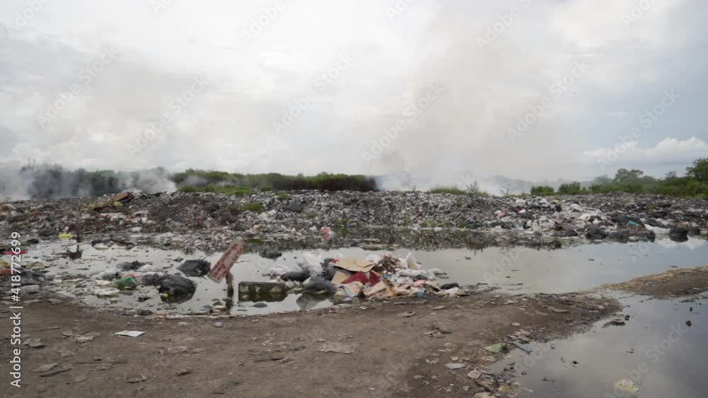 Pan of trash dump with smoldering garbage, hazy sky, and pools of ...