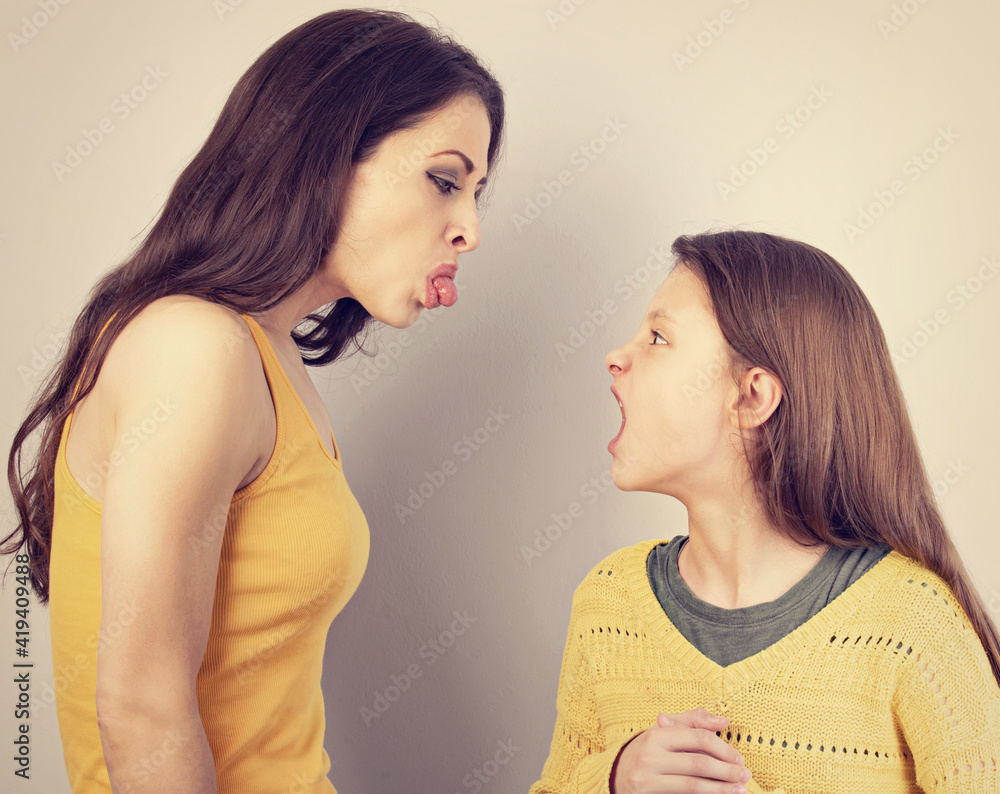 Angry strict mother showing the tongue and her emotional anger daughter ...