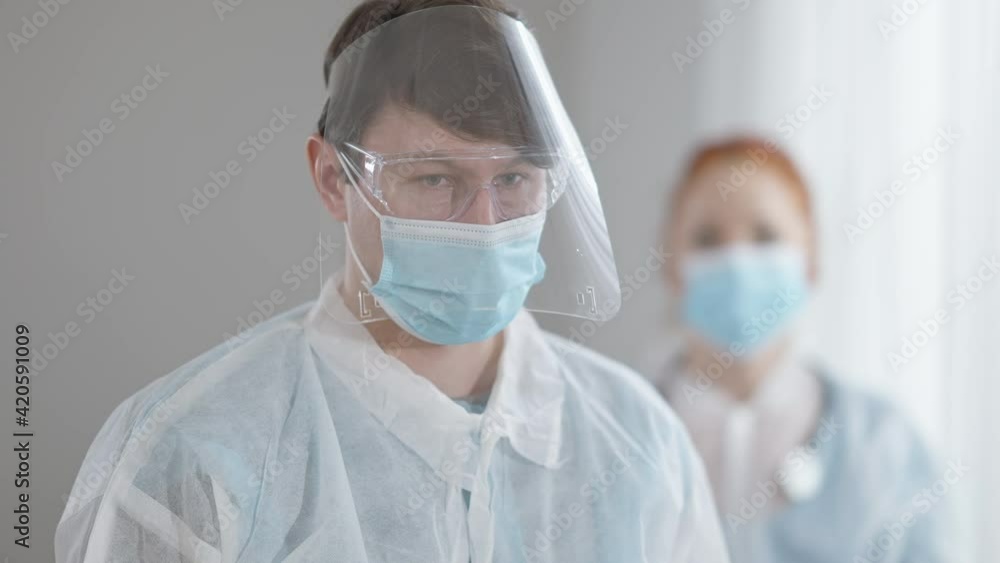 Concentrated male doctor putting on face shield with blurred female ...
