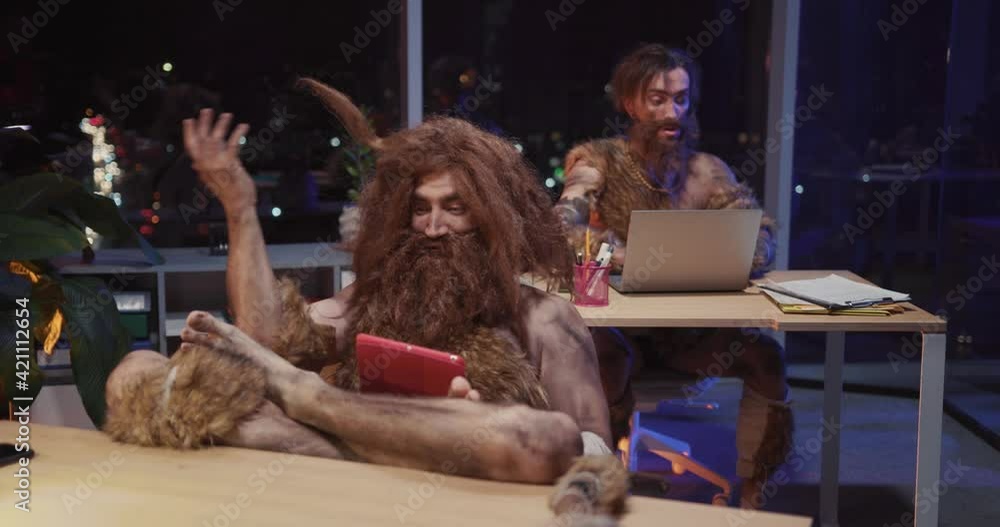 Bearded prehistoric caveman in fur clothes sitting on office desk using ...