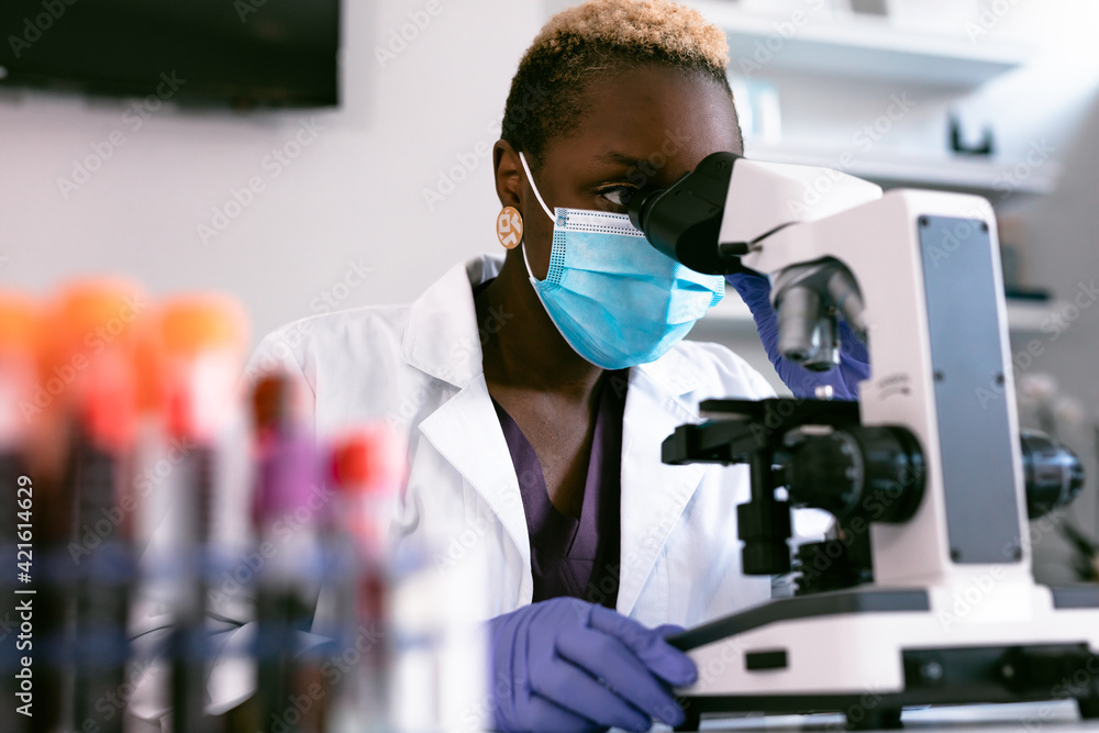 Black female scientist in mask in laboratory looking through microscope ...