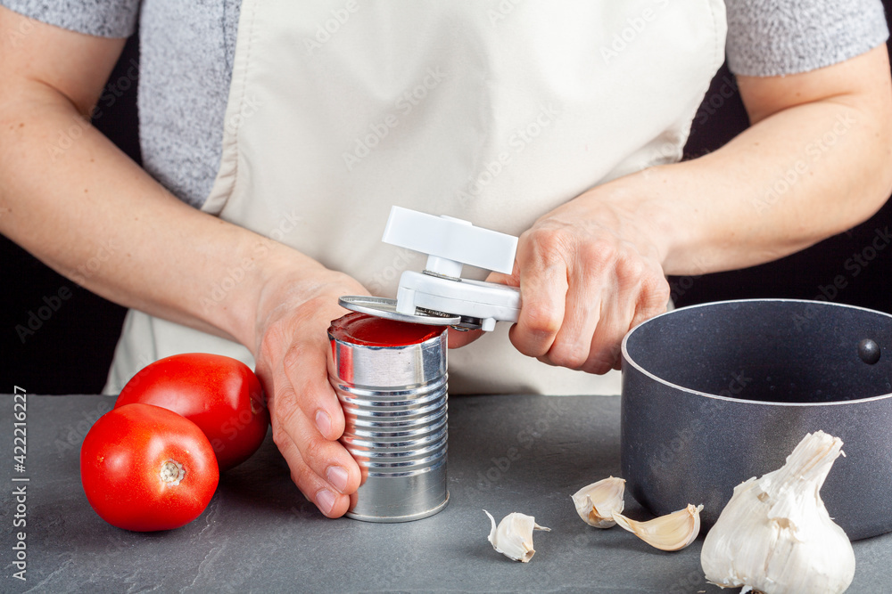 A woman is carefully opening a can of tomato paste on a kitchen counter ...