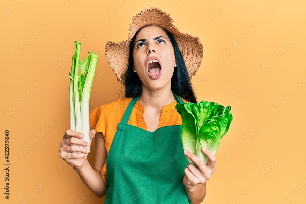 Beautiful young woman wearing gardener apron holding vegetables angry ...
