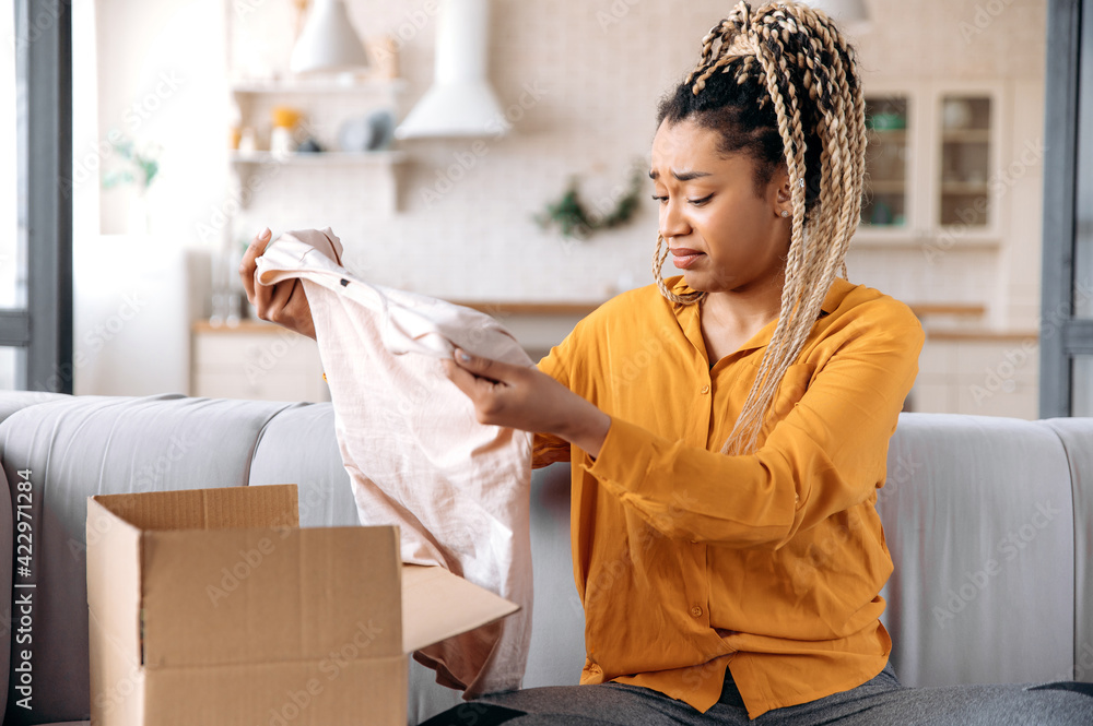 A confused sad african american girl unpacking her parcel at home ...
