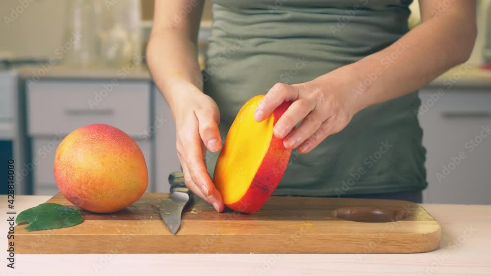 Woman cuts mangoes on table of house in kitchen. Hands hold mango and ...