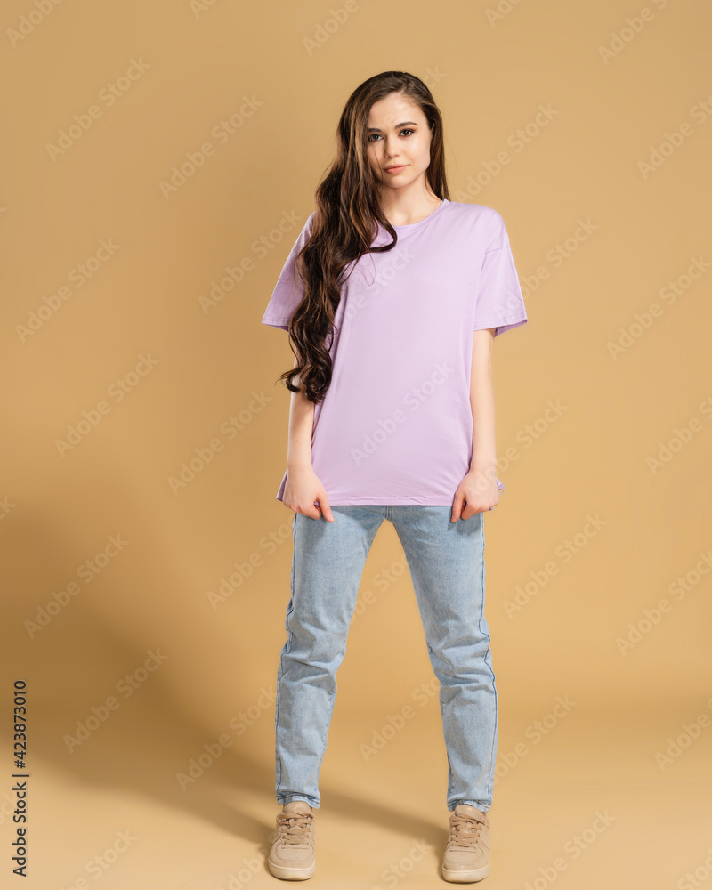 Young beautiful girl with long curly hair in a lilac T-shirt and blue jeans on a pastel orange ...
