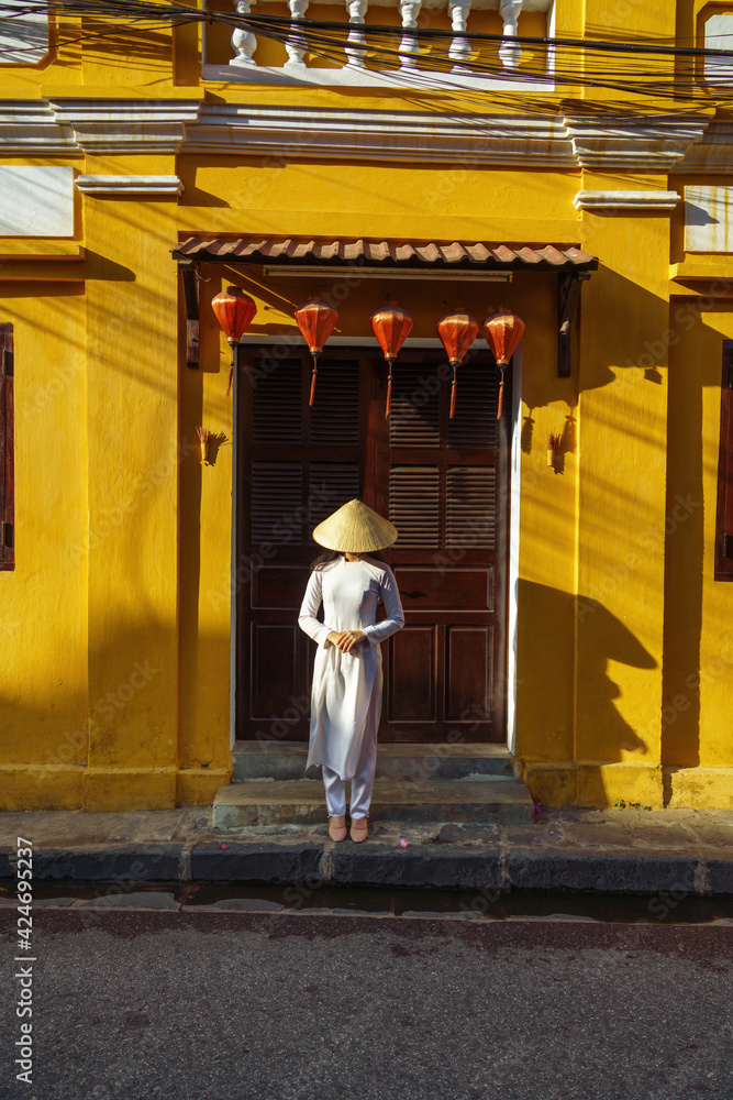 A young girl in traditional dress Ao Dai and conical hat standing on Hoi An ancient street Stock ...