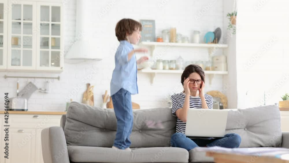 Irritated Mother Sitting On Couch Working On Laptop At Home Distracted