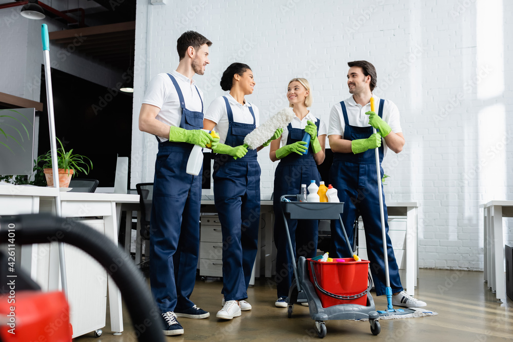 Smiling multiethnic cleaners talking near cart with detergents in ...
