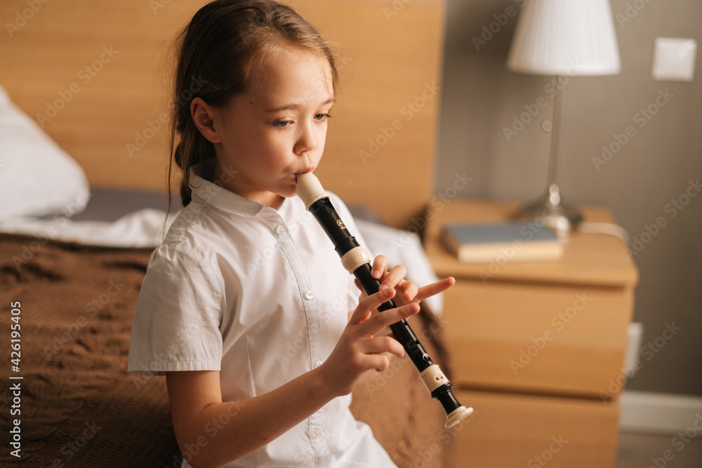 Side view of talented adorable little girl playing flute sitting on bed ...