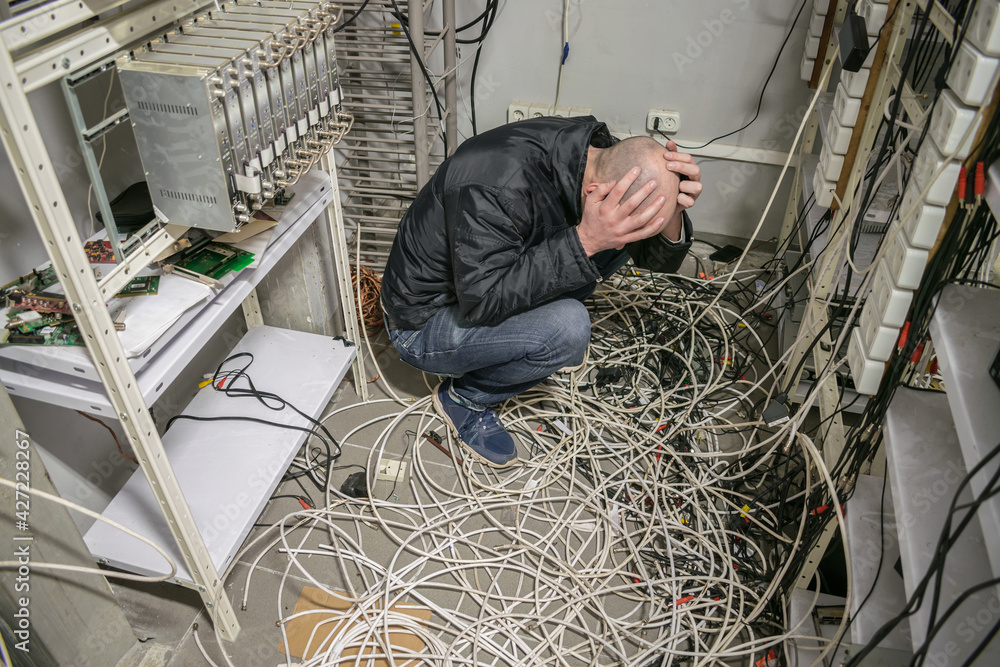 A sad technician sits near a pile of wires and holds his head with his ...