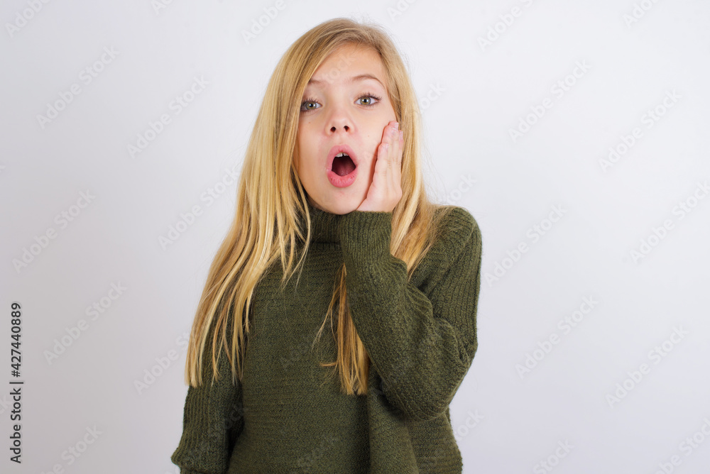 Shocked Caucasian kid girl wearing green knitted sweater against white ...
