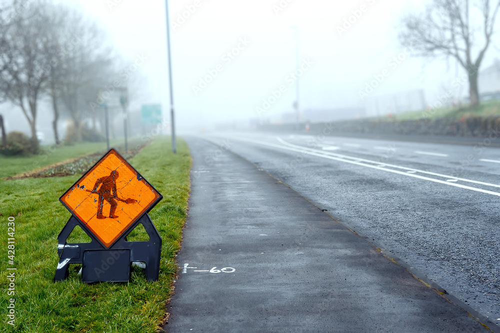 Fotka „Road work sign in focus. Asphalt road in town in fog. Dangerous ...