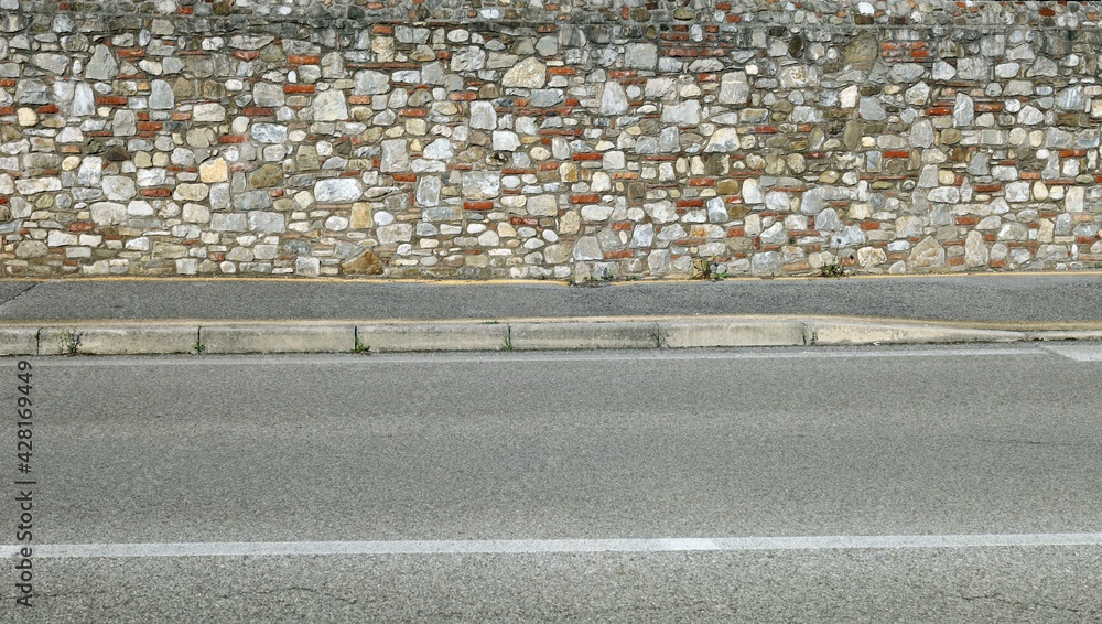Rustic stone wall made of rocks and bricks. A sidewalk and asphalt road ...