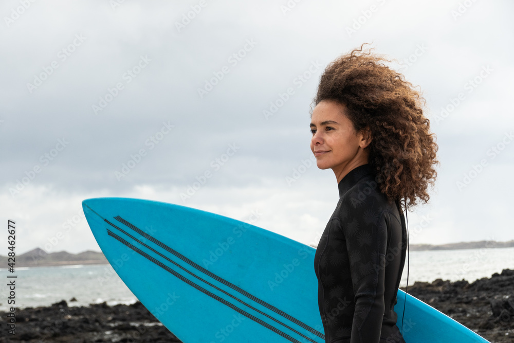 Afro surfer girl wearing wetsuit standing at rocky surf spot in a ...