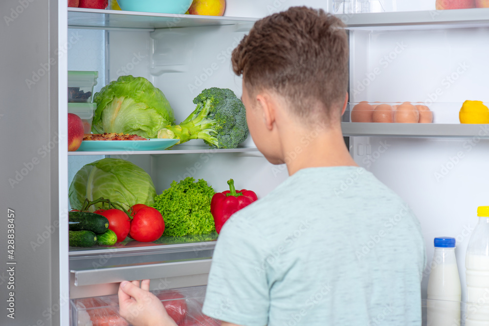 Back view - young teen boy standing near open fridge in kitchen at home ...