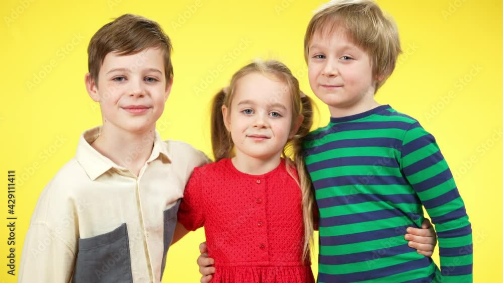 Three charming children hugging at yellow background and smiling looking at camera. Portrait of ...