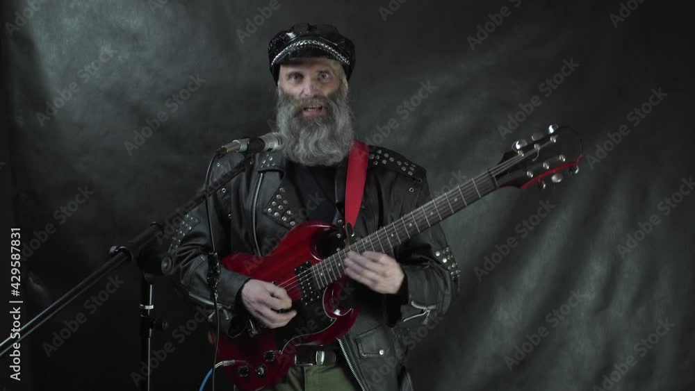 Bearded rock musician in leather jacket and cap plays red electric ...