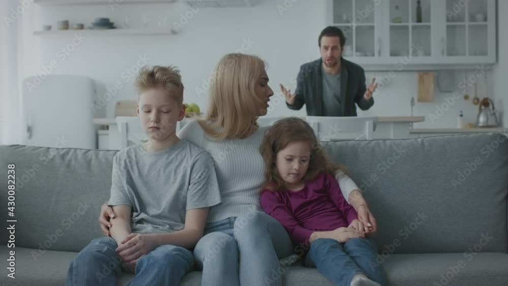 Unhappy mother sitting on sofa embracing sad kids and crying, angry man ...