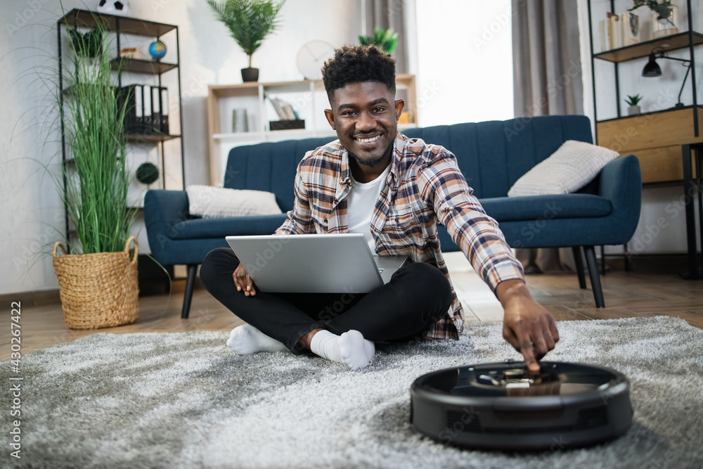 Smiling african man turning on robot vacuum cleaner while for cleaning ...