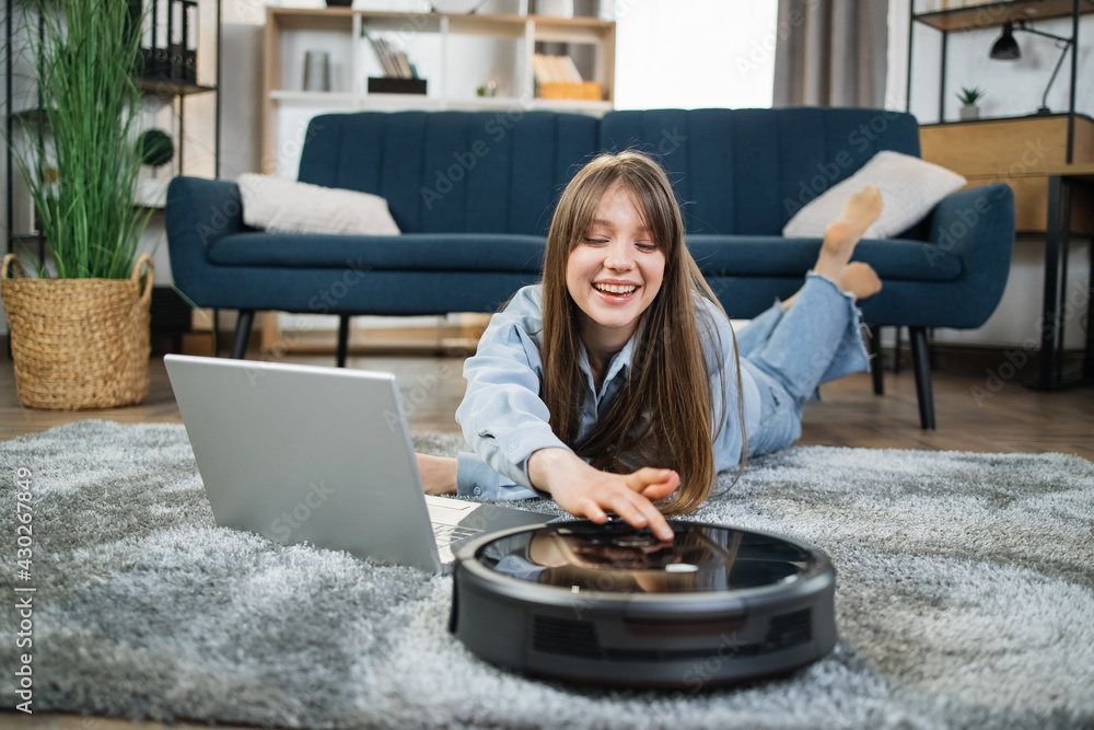 Happy young woman in casual wear using robot vacuum cleaner for chores ...