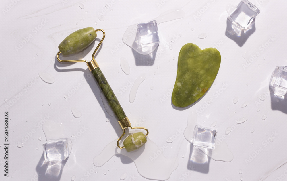 A gua sha roller and a jade stone scraper and ice on a white background ...