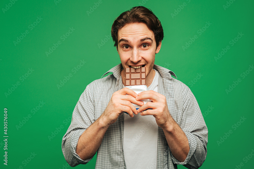 Young man enjoys eating of chocolate bar, isolated on green background ...