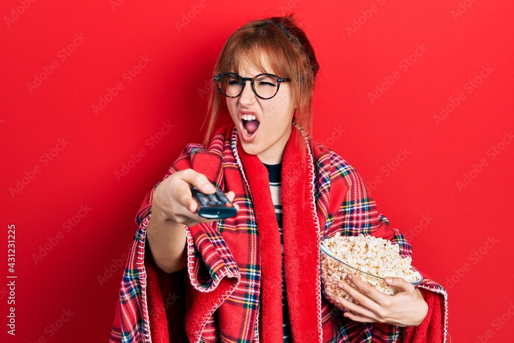 Redhead young woman eating popcorn using tv control angry and mad ...