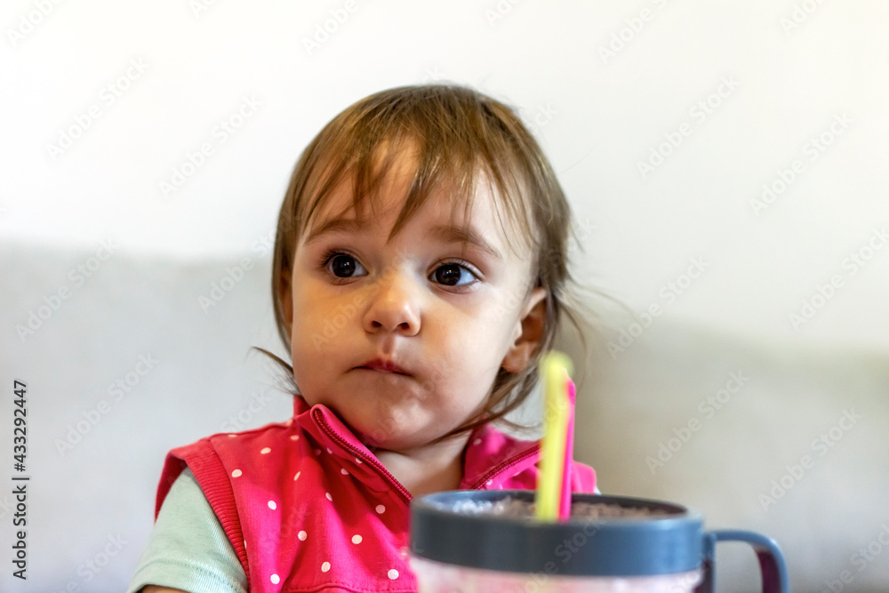 Photo of cute baby girl holding plastic glass with straw while drinking ...