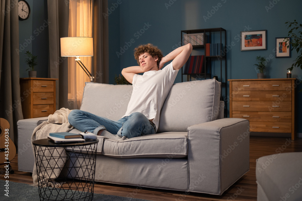 A young boy sits on the living room couch in the evening with his legs ...