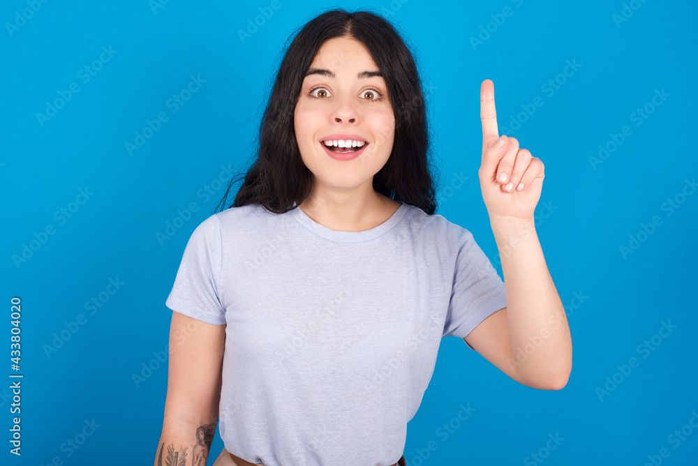 young beautiful tattooed girl wearing blue t-shirt standing against blue background pointing ...