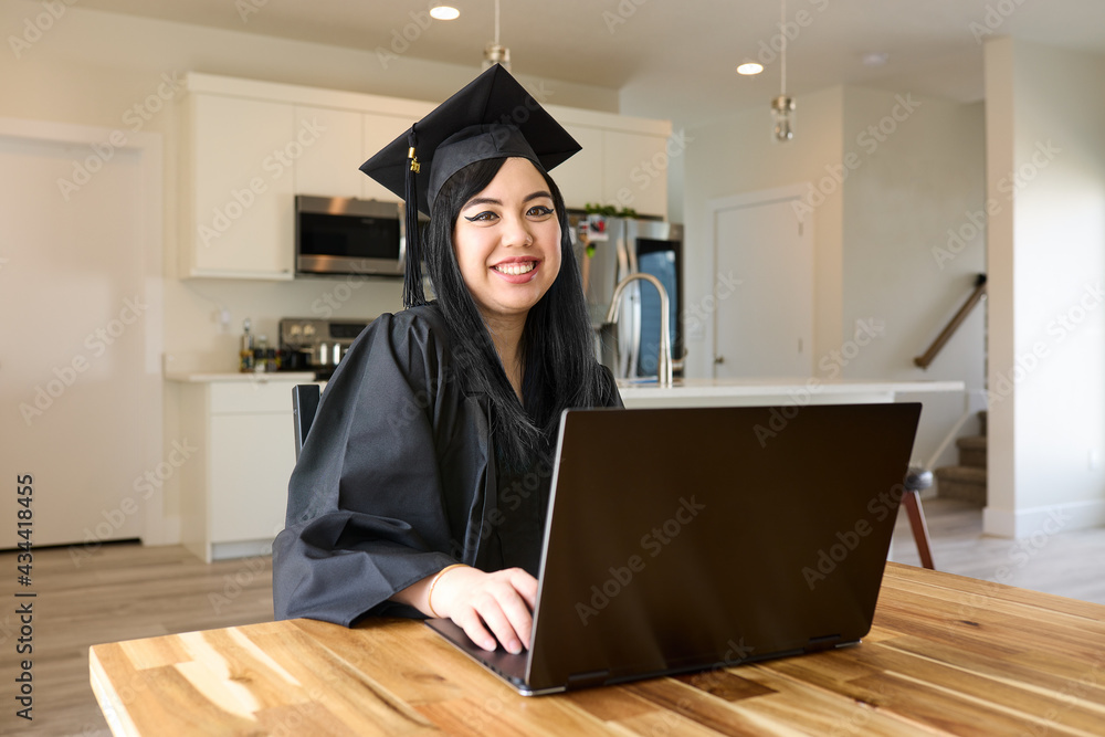 Smiling Asian woman in graduation gown sitting in kitchen at table with ...