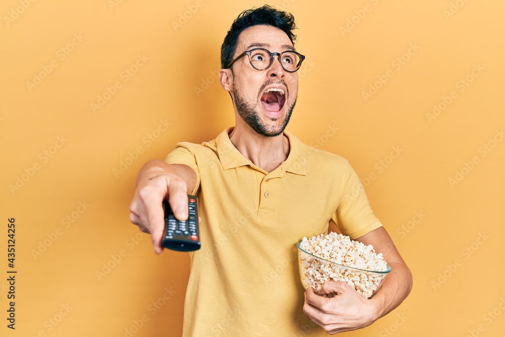 Young hispanic man eating popcorn using tv control angry and mad ...